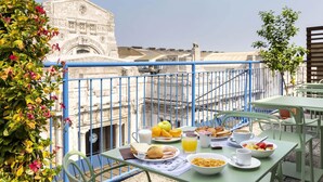 Varanda com mesa posta para o café da manhã, incluindo pão, frutas e bebidas, com vista para um edifício histórico de arquitetura detalhada.