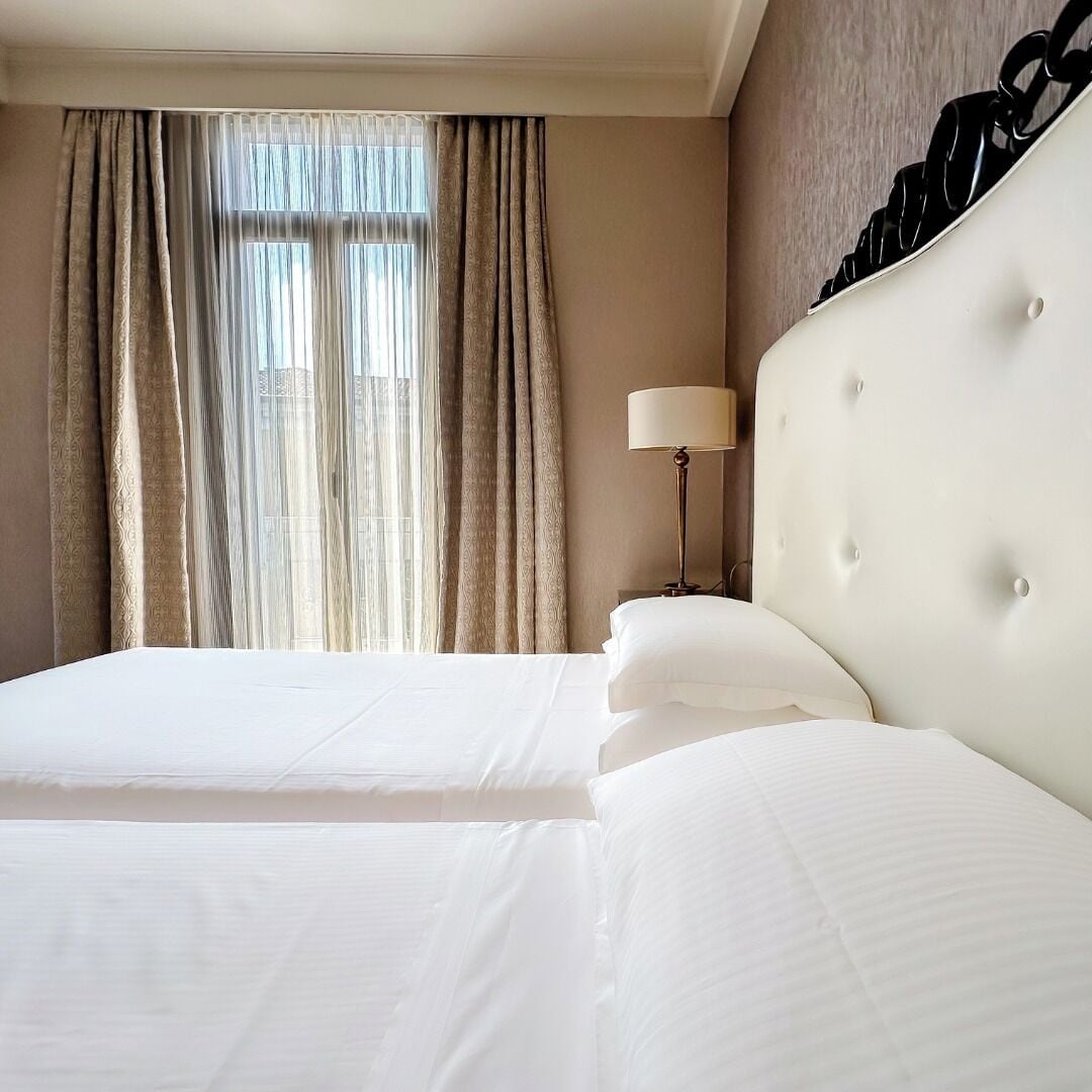 Quarto de hotel com uma cama grande, abajur e vista através de cortinas leves.