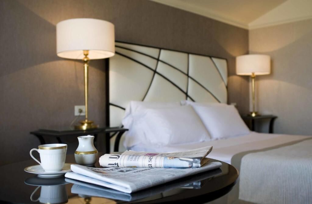 Quarto de hotel com uma cama bem arrumada, duas luminárias de cabeceira, uma mesinha com uma xícara, um jarro e jornais.