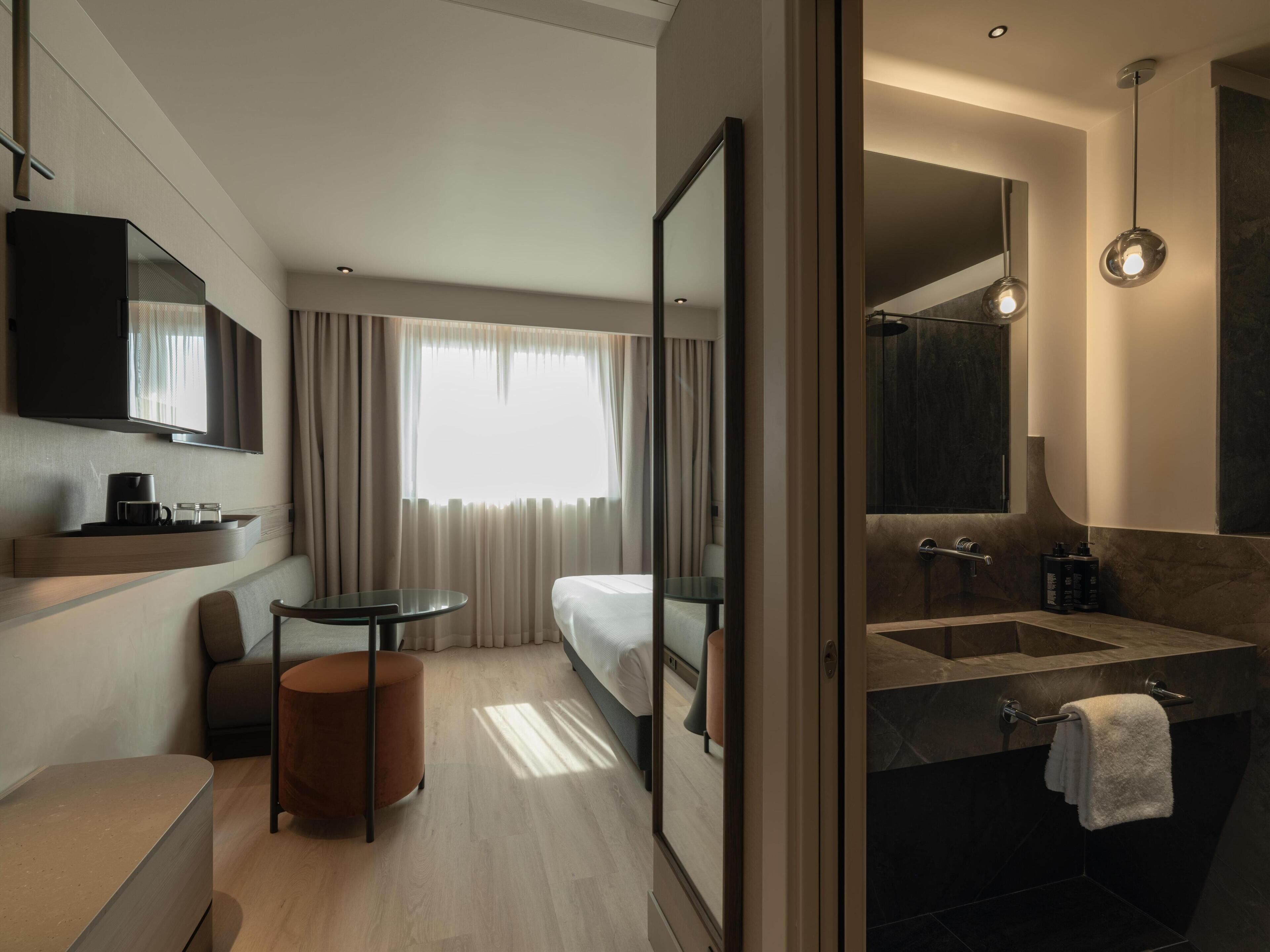 Quarto de hotel moderno com cama, escrivaninha, cadeira, um banquinho redondo, televisão, um espelho grande e banheiro com pia e porta-toalhas.