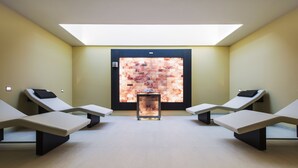 Um espaço de spa com duas cadeiras reclináveis, uma mesa de massagem central e uma grande janela com vista para uma parede de tijolos.