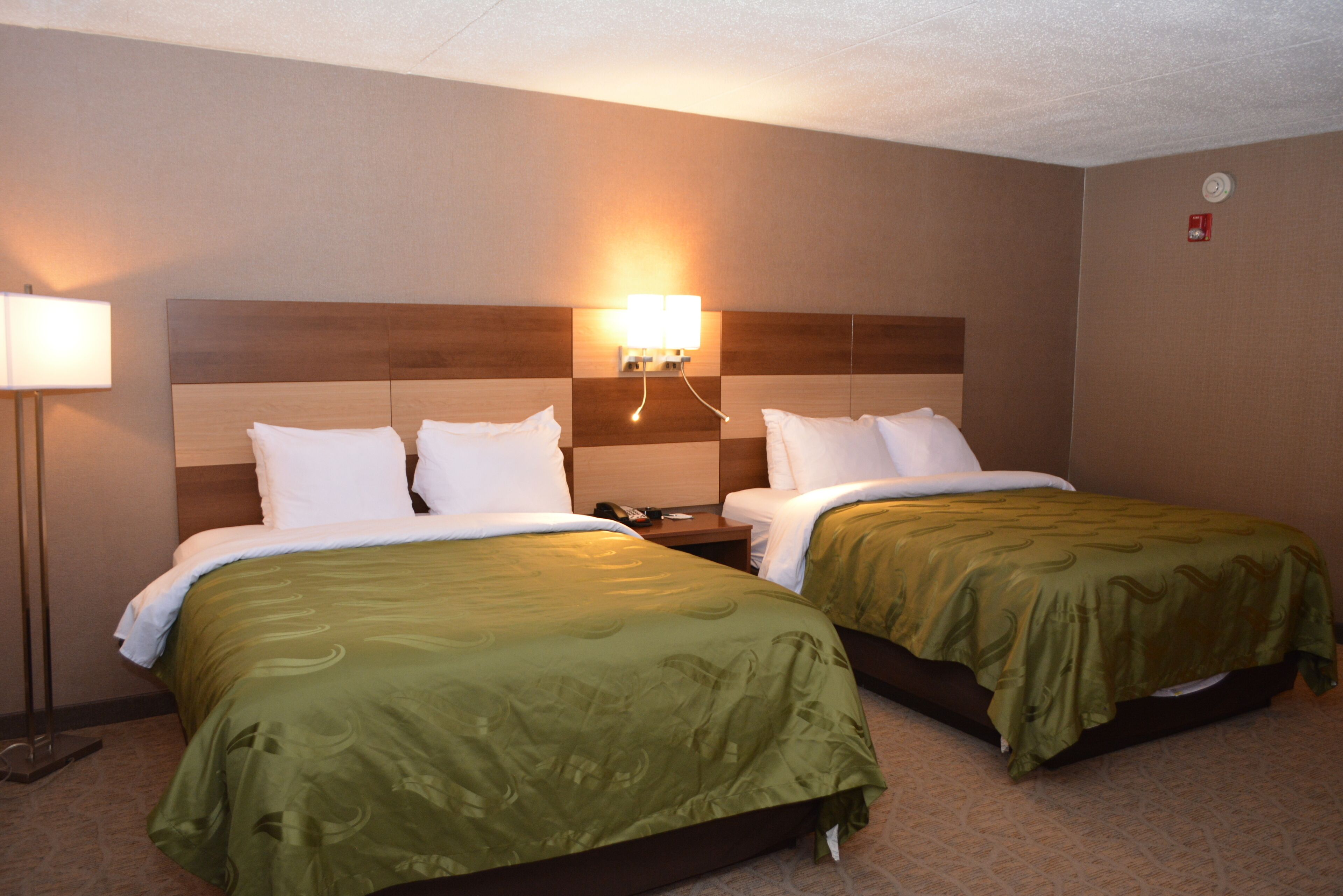 Quarto de hotel com duas camas, cada uma com colcha verde, mesa de cabeceira com abajur e luminária na parede.