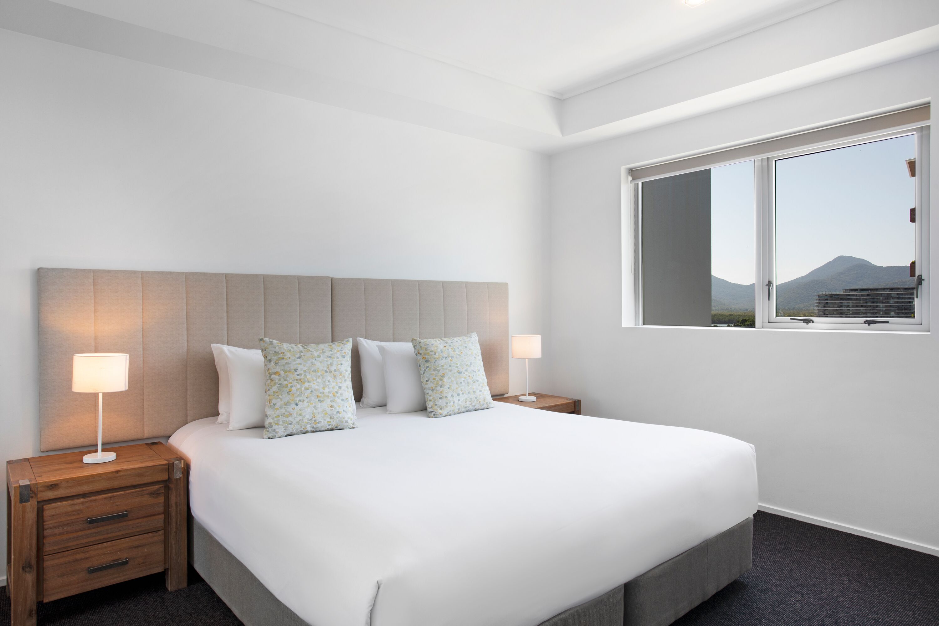 Uma cama bem arrumada com cabeceira, duas mesas de cabeceira com abajures e uma janela com vista para montanhas.