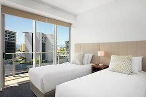 Quarto de hotel com duas camas, uma janela ampla com vista para a cidade e uma mesa de cabeceira com abajur.