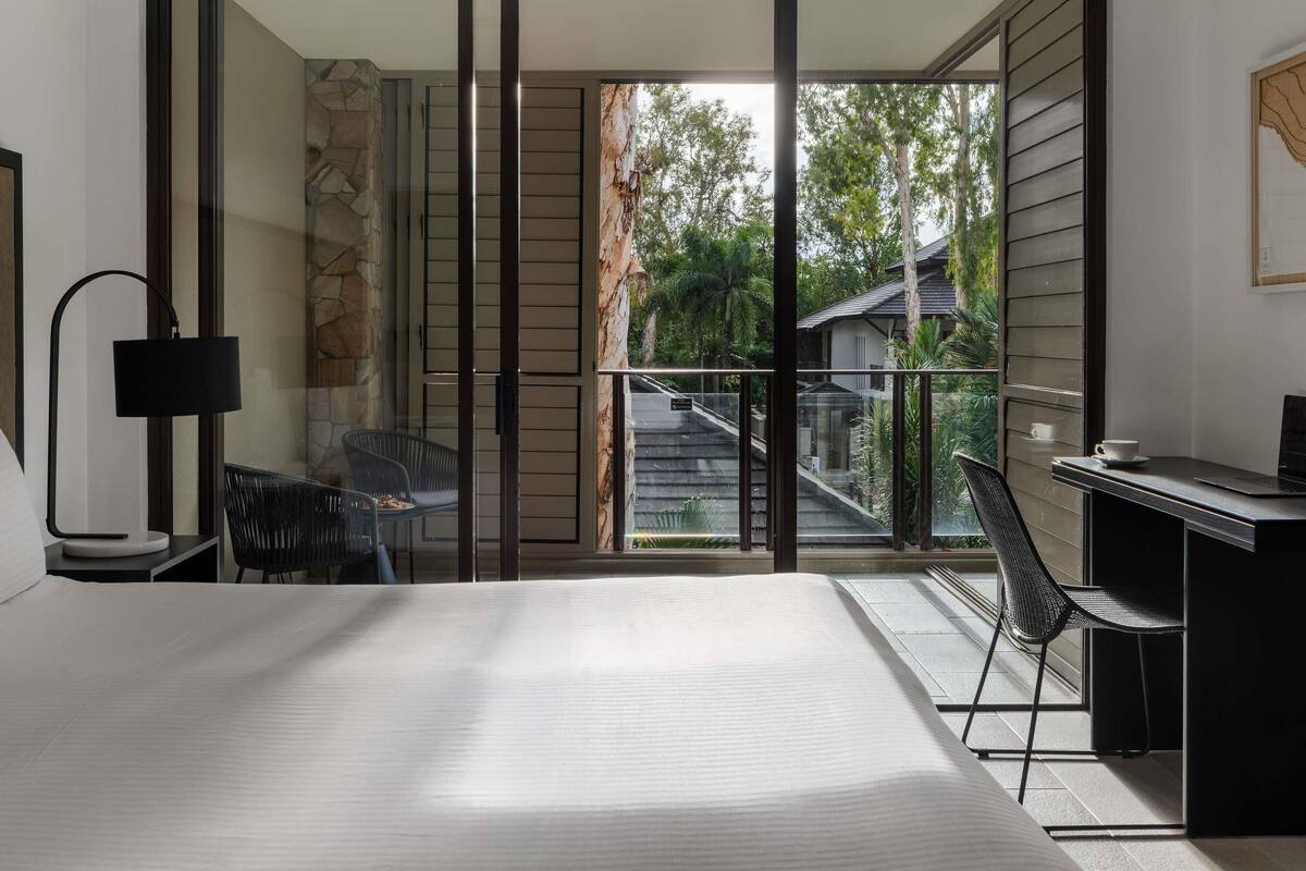 Quarto de hotel moderno com uma cama grande, uma escrivaninha com um laptop, uma cadeira e uma varanda com vista para árvores e uma escadaria.