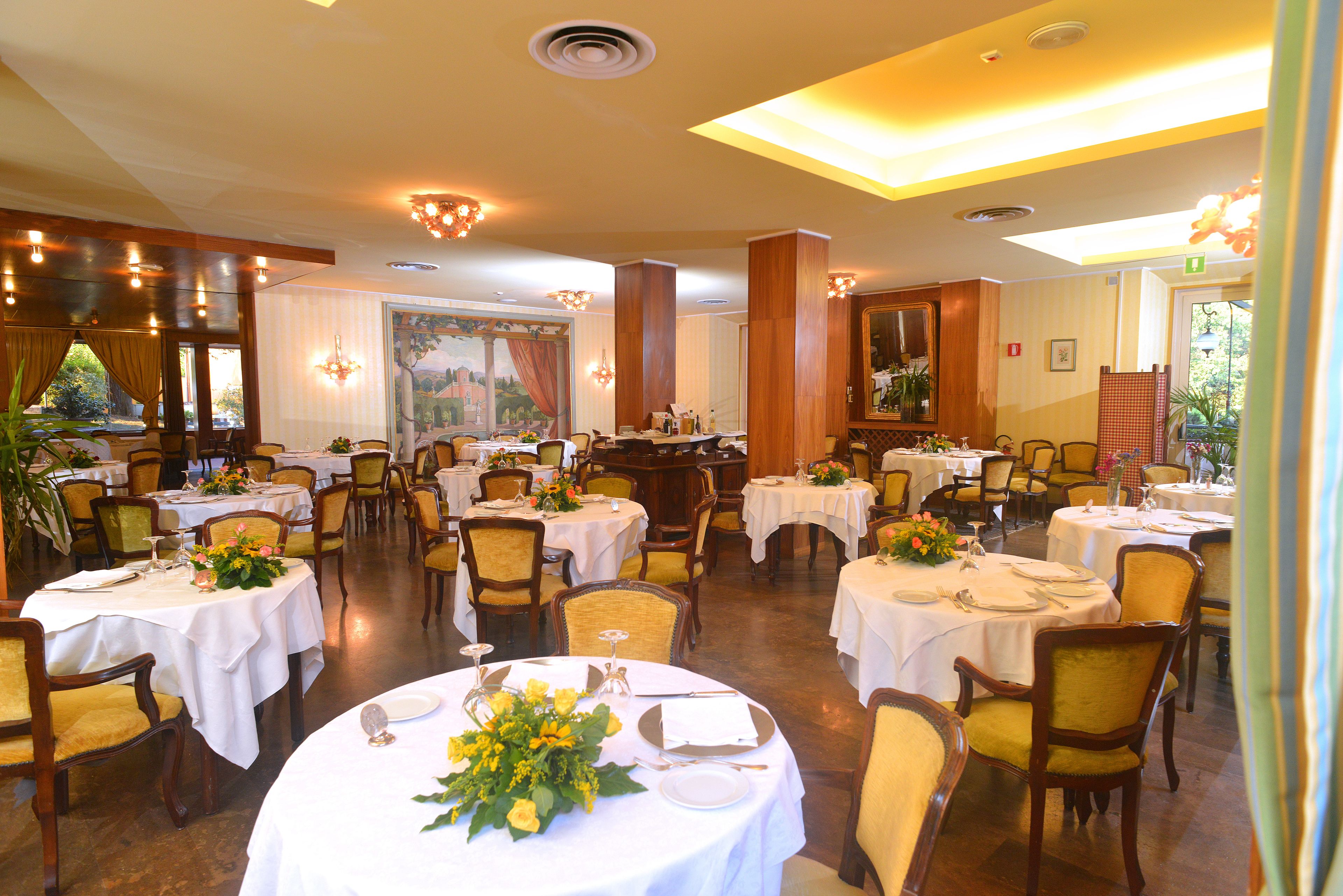 Uma ampla sala de jantar com várias mesas redondas preparadas para refeições, cada uma com toalhas brancas e centros de mesa florais.