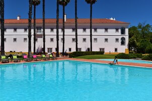 Um edifício grande e branco com piscina na frente, cercado por palmeiras e espreguiçadeiras.