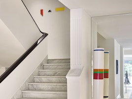 Uma escadaria com degraus de mármore e corrimão preto, que leva a um patamar com parede branca e radiador.