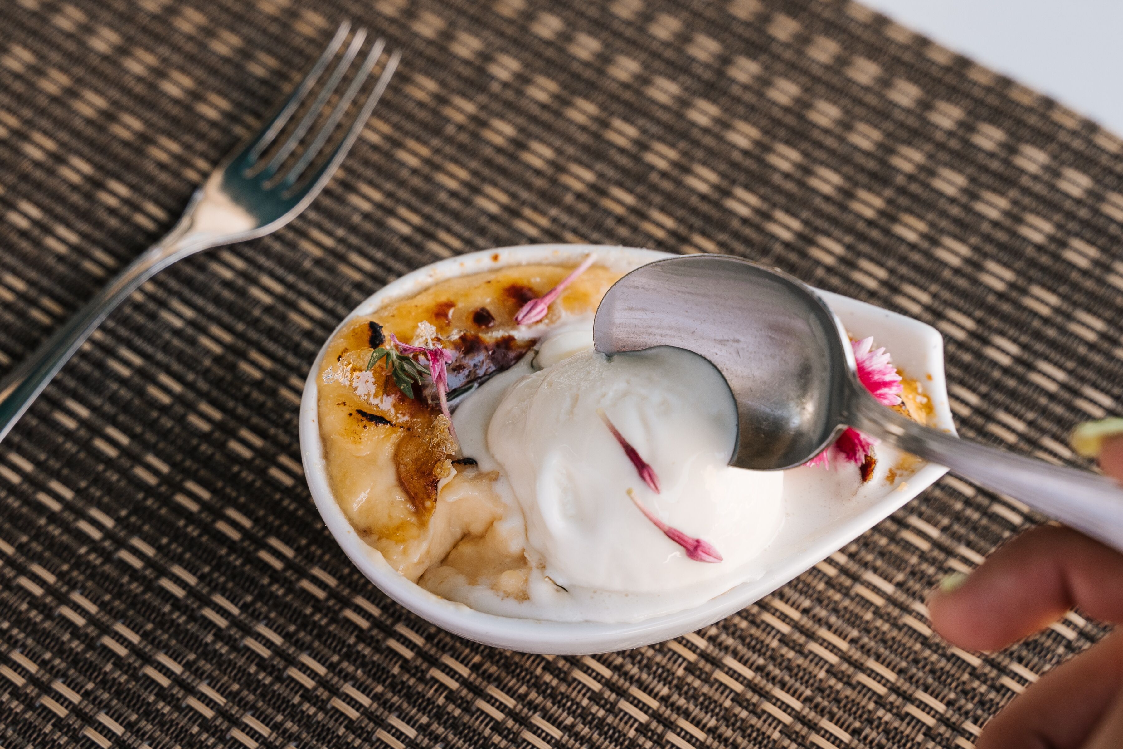 Uma tigela branca com uma colher, contendo uma sobremesa cremosa com um toque de chantilly e um enfeite de flor rosa.