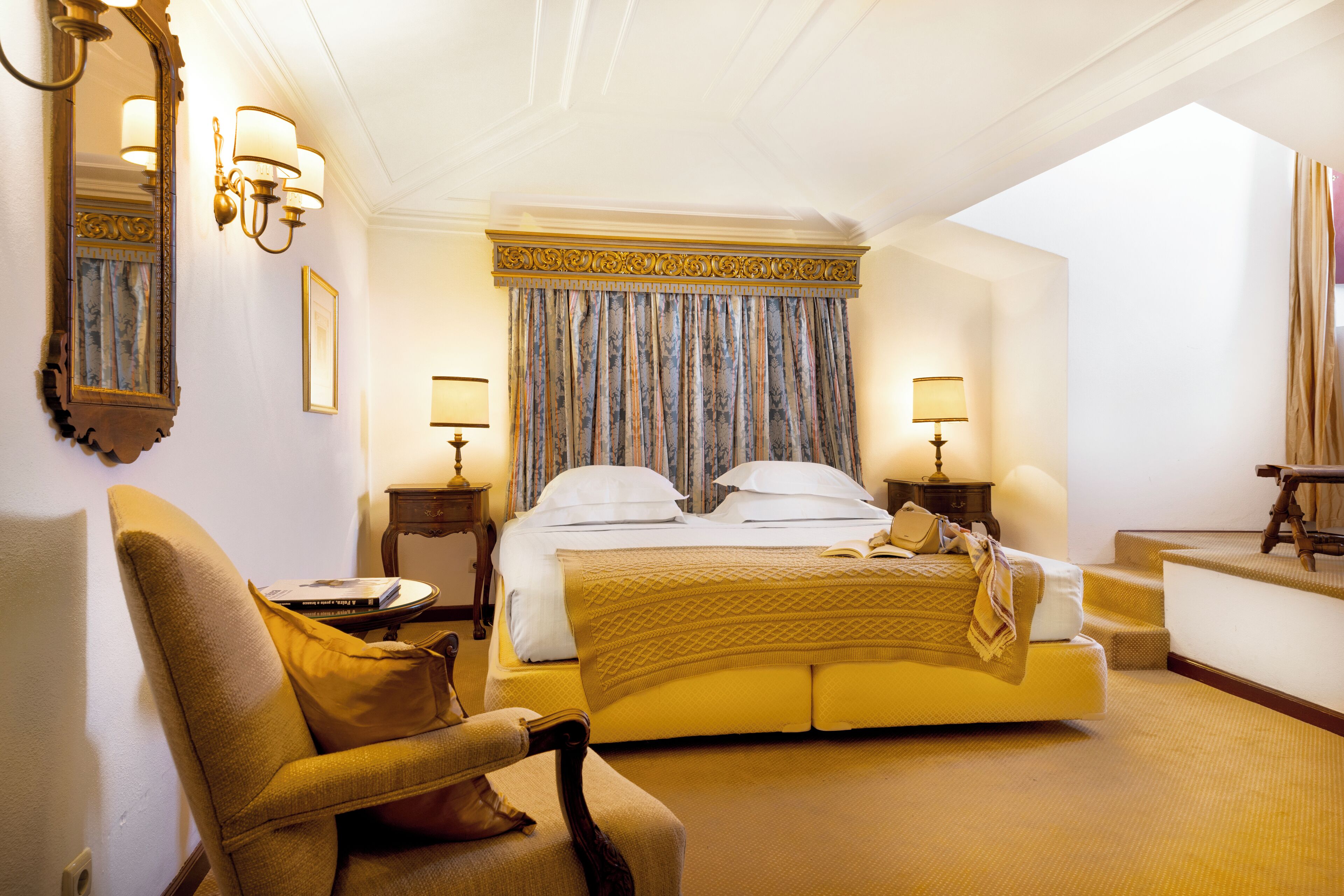 Quarto de hotel com uma cama grande, duas mesinhas de cabeceira, uma cadeira, um espelho e uma janela com cortinas.
