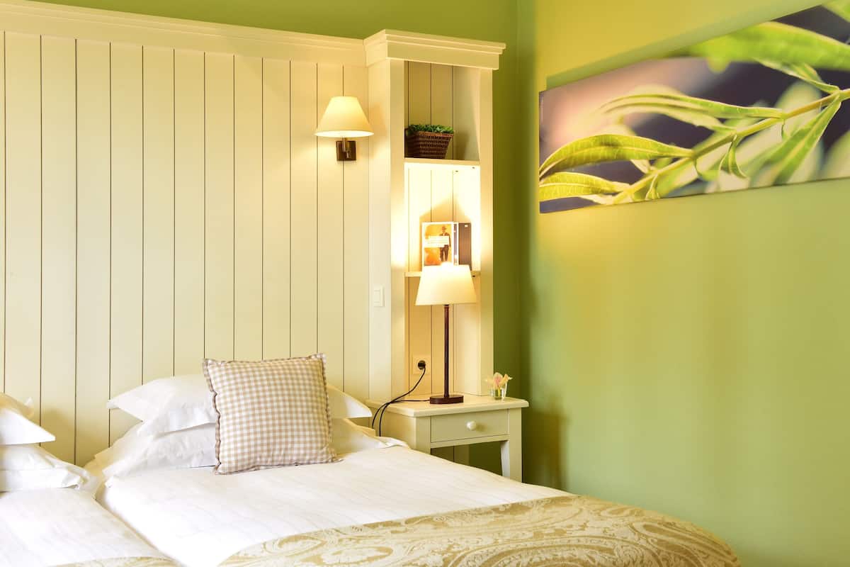 Uma cama bem arrumada com travesseiro xadrez, luminária de cabeceira e mesa de apoio com um pequeno vaso de flores.
