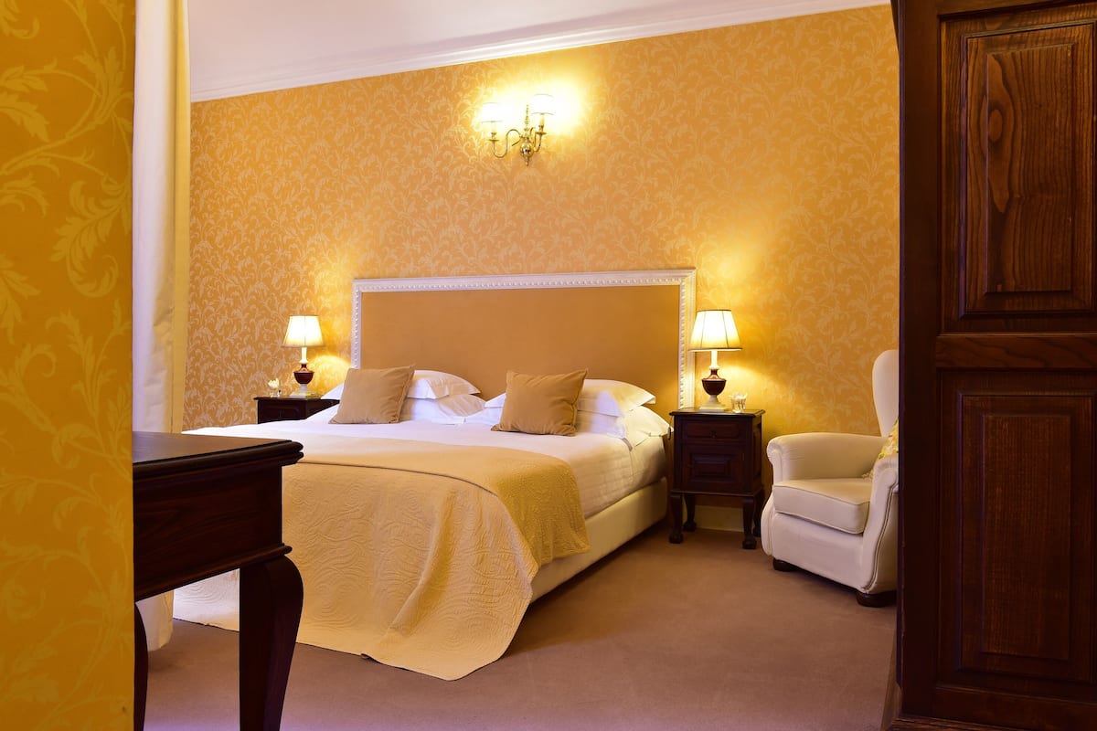 Quarto de hotel com uma cama grande, duas mesas de cabeceira com luminárias, uma cadeira e um guarda-roupa de madeira.