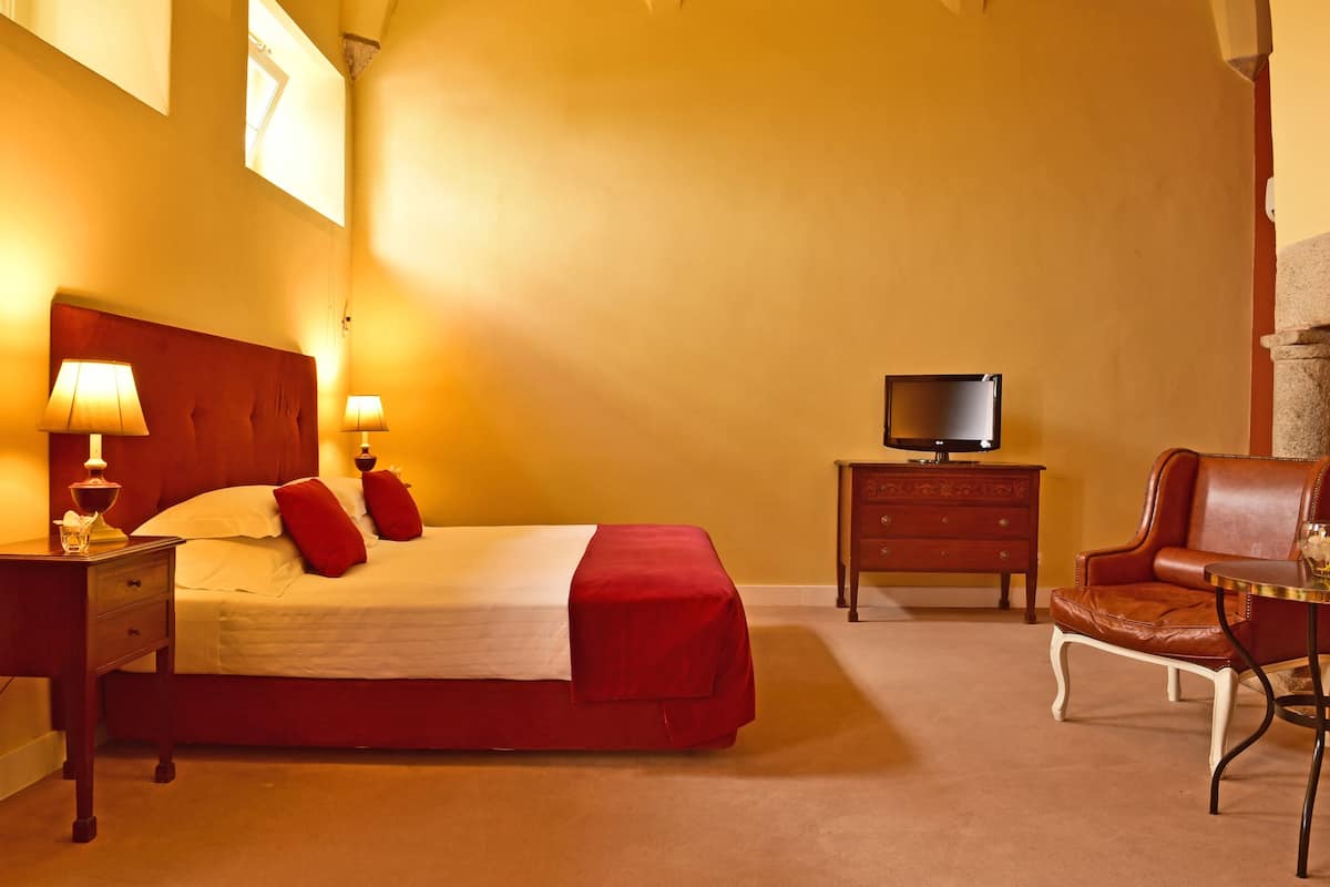 Quarto de hotel com uma cama grande, duas luminárias de cabeceira, uma televisão sobre uma mesa de cabeceira de madeira, uma poltrona de couro e uma mesinha.