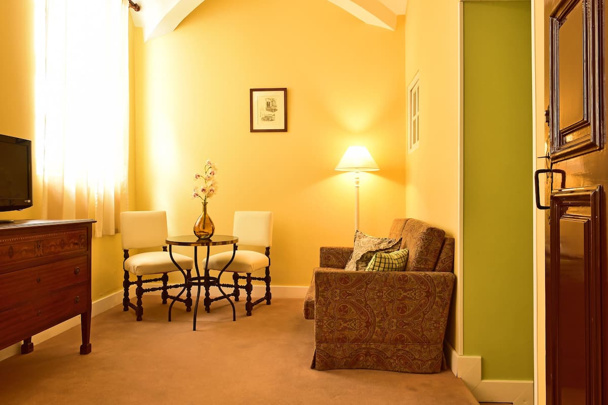 Um quarto aconchegante com penteadeira de madeira, uma mesinha redonda com cadeiras, uma poltrona estampada e um abajur.