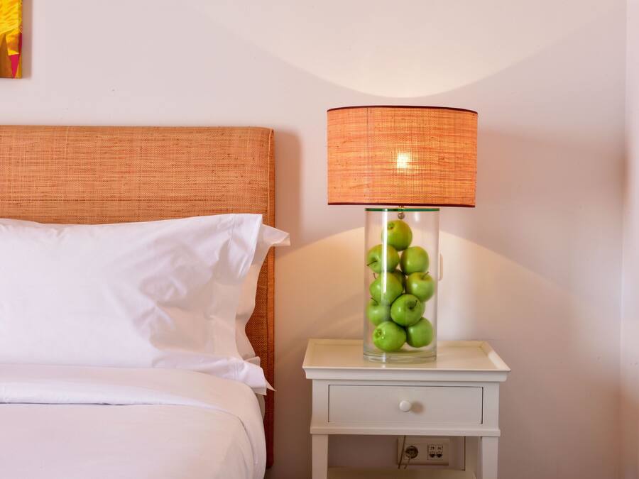Quarto de hotel com cama, roupa de cama branca, mesa de cabeceira com abajur e um frasco de vidro com maçãs verdes.