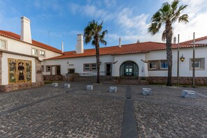 Um pátio com piso de lajes, palmeiras e um edifício com telhado de telhas vermelhas e paredes brancas.