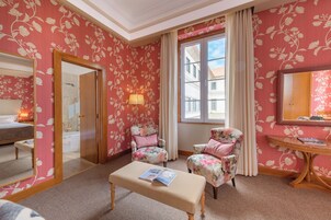 Quarto com papel de parede floral, uma cama, um banco com uma revista, duas poltronas florais, uma mesa com um abajur e uma janela grande.