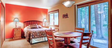 Um quarto com cama, mesas de cabeceira, uma mesa de jantar de madeira com cadeiras, um candelabro e uma janela grande com vista para árvores.
