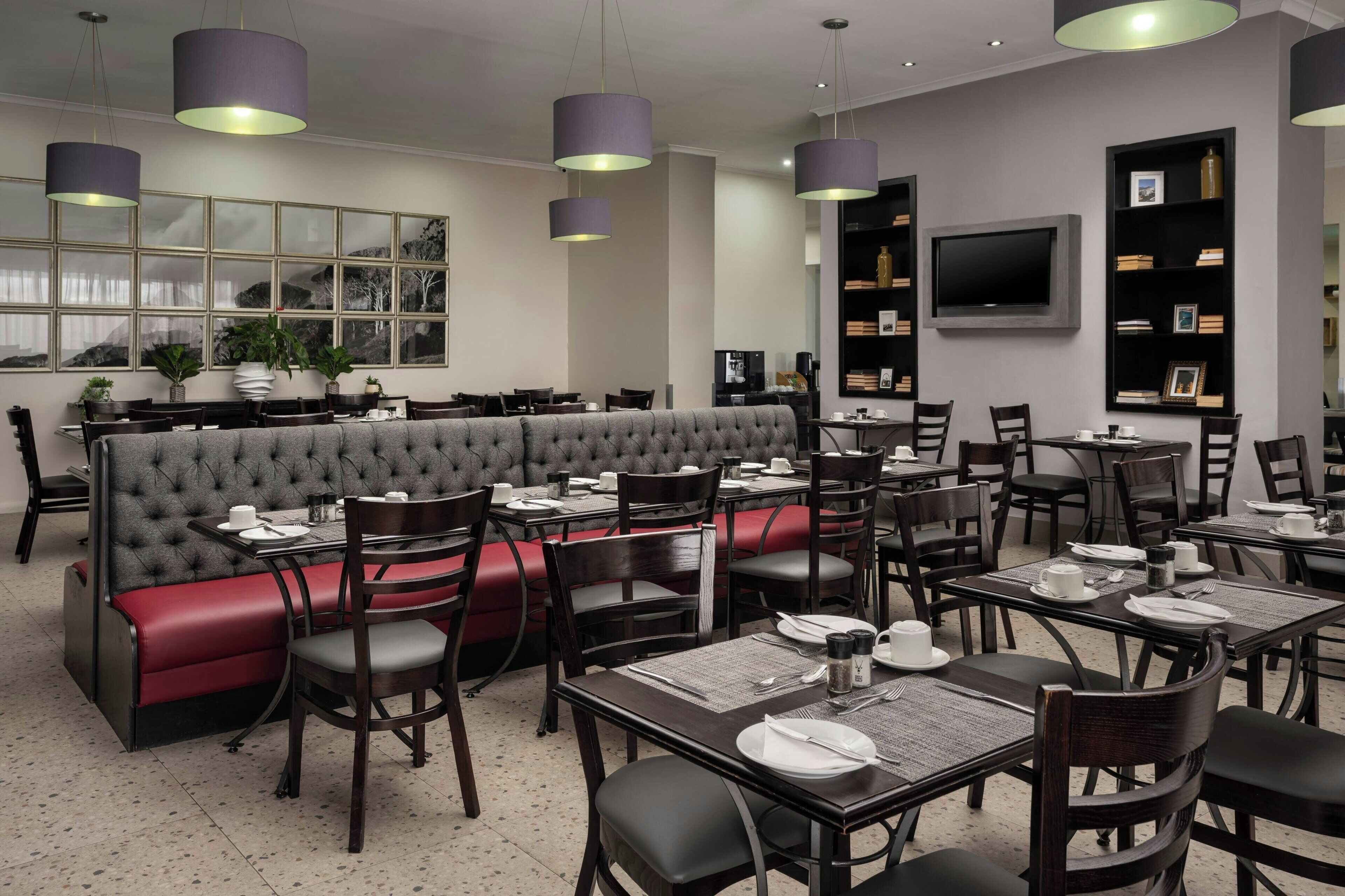 Um restaurante com assentos em cabine estofados em cinza, mesas e cadeiras de madeira escura. Há luminárias pendentes no teto, uma televisão de tela plana na parede e uma estante com diversos objetos.