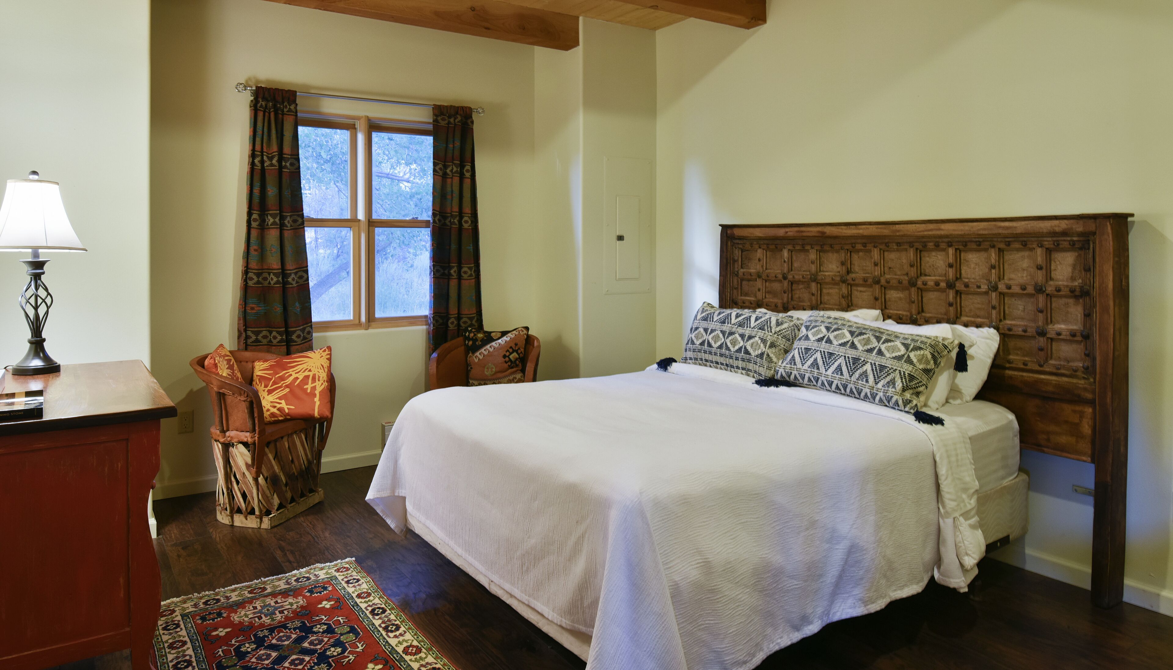 Um quarto com cabeceira de madeira, uma cama com almofadas estampadas, uma cadeira de madeira, uma escrivaninha com abajur e uma janela com cortinas.