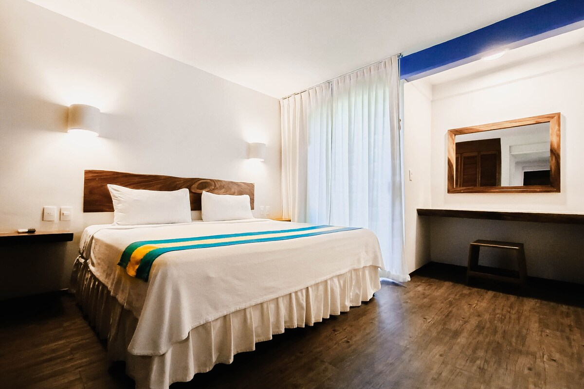 Quarto de hotel com cama, mesas de cabeceira, espelho e piso de madeira.
