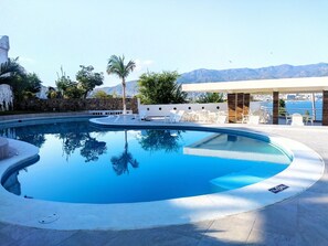 Uma piscina com borda curva, rodeada por espreguiçadeiras brancas e um muro de pedra.