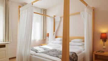 Uma cama de dossel com lençóis brancos, uma mesa de cabeceira com abajur e uma janela com cortinas.