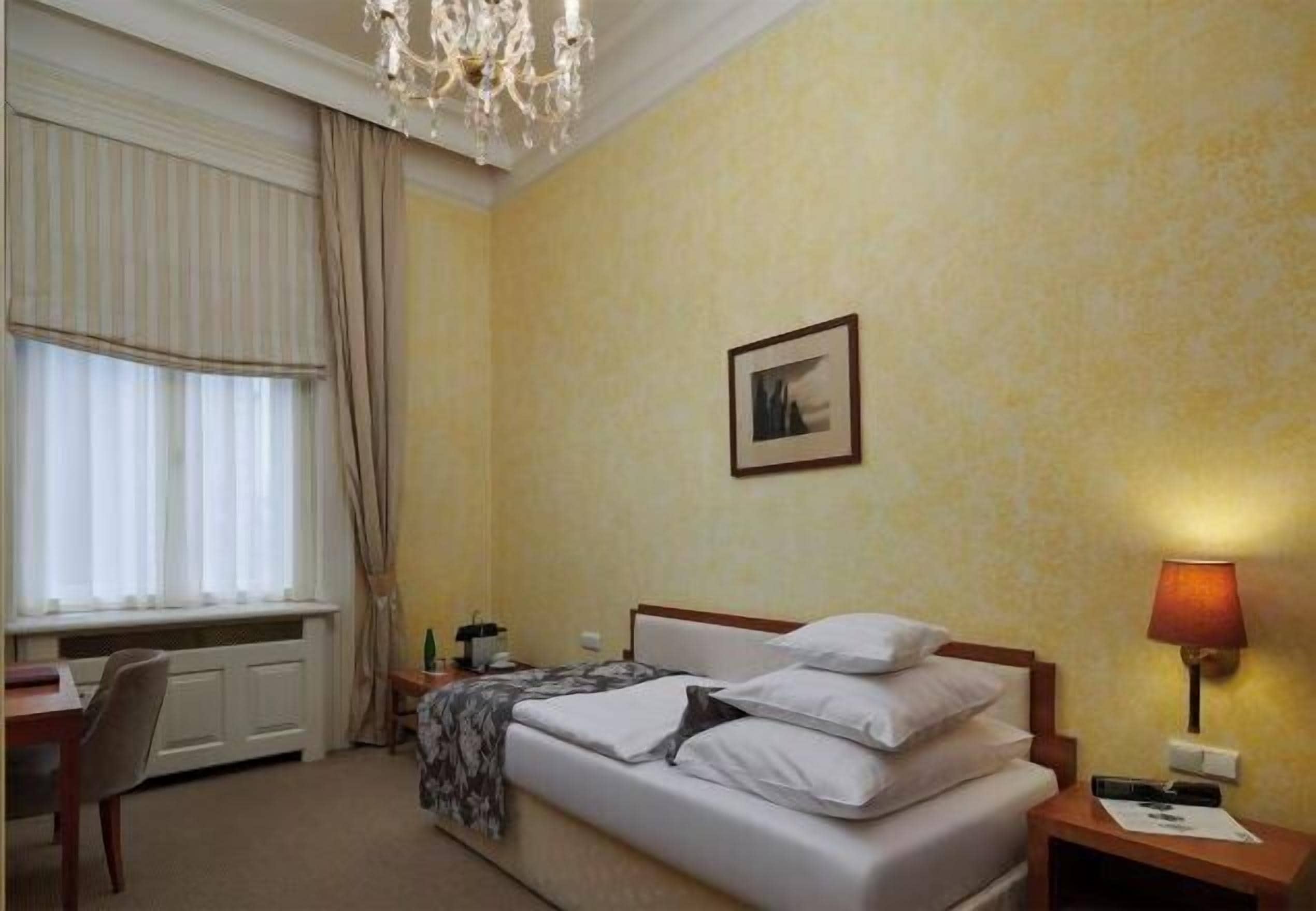 Quarto de hotel com cama, escrivaninha, cadeira, abajur e um quadro na parede.