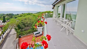 Uma varanda com vista para o mar, cadeiras brancas e um carro estacionado na rua.