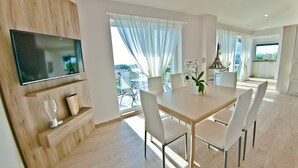 Uma sala de jantar moderna com uma mesa grande, cadeiras brancas e uma televisão de tela plana fixada numa parede de madeira.