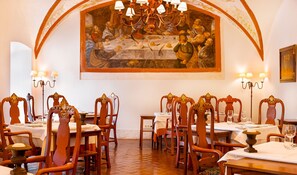 Uma área de refeições com cadeiras de madeira ricamente decoradas, mesas com toalhas brancas e um quadro grande retratando uma cena histórica acima das mesas.