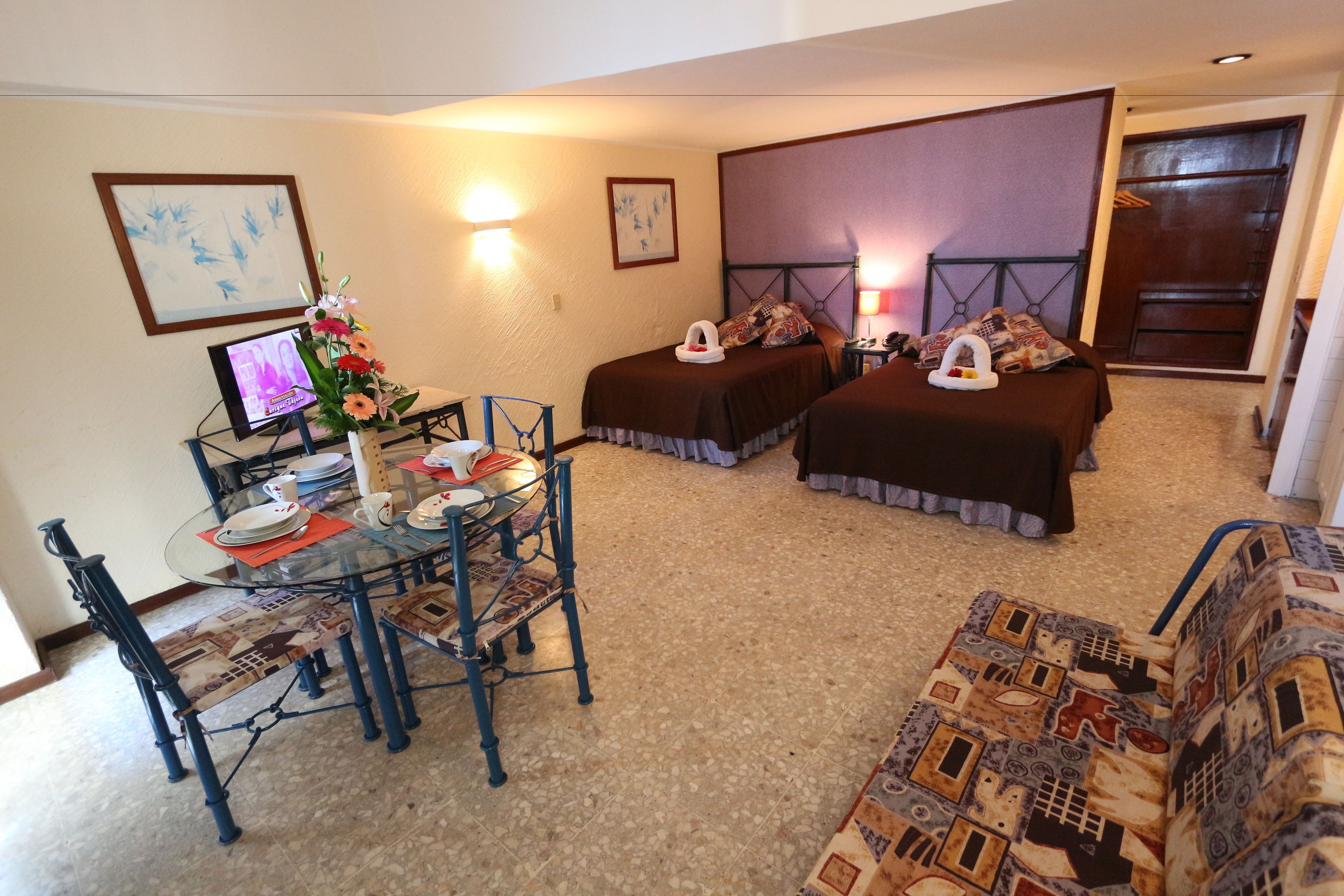 Quarto de hotel com duas camas, uma mesa de jantar com cadeiras, uma televisão pequena e um vaso decorativo.