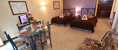 Quarto de hotel com duas camas, uma mesa de jantar com cadeiras, uma televisão pequena e um vaso decorativo.