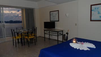 Quarto de hotel com varanda, cama com cobertura azul, mesa com cadeiras, televisão e uma garrafa de champanhe.