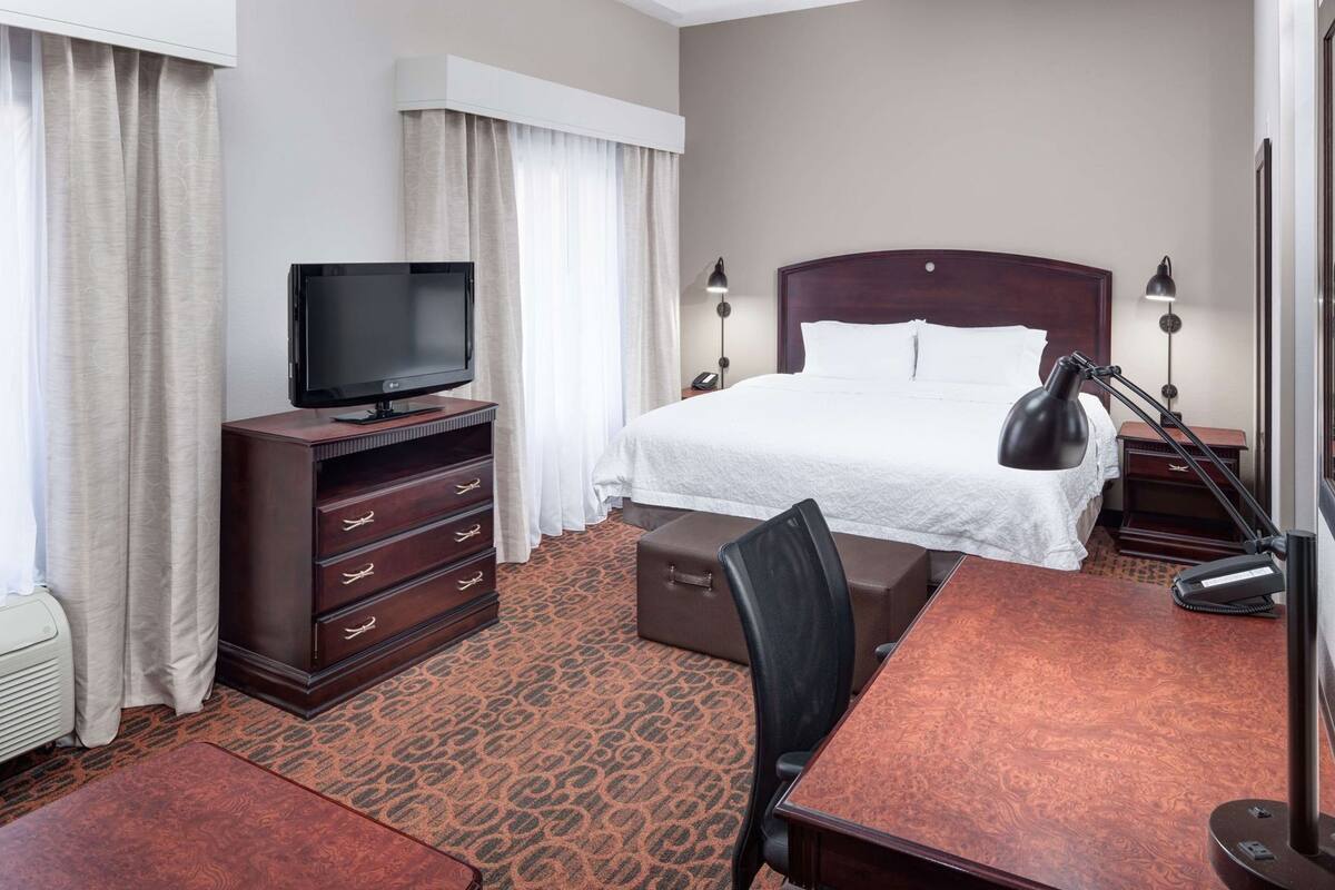 Quarto de hotel com cama, escrivaninha, televisão, luminária de mesa, cadeira e aquecedor.