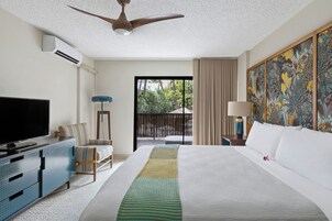 Quarto de hotel com cama, televisão, escrivaninha, cadeira, varanda com cortinas e ventilador de teto.