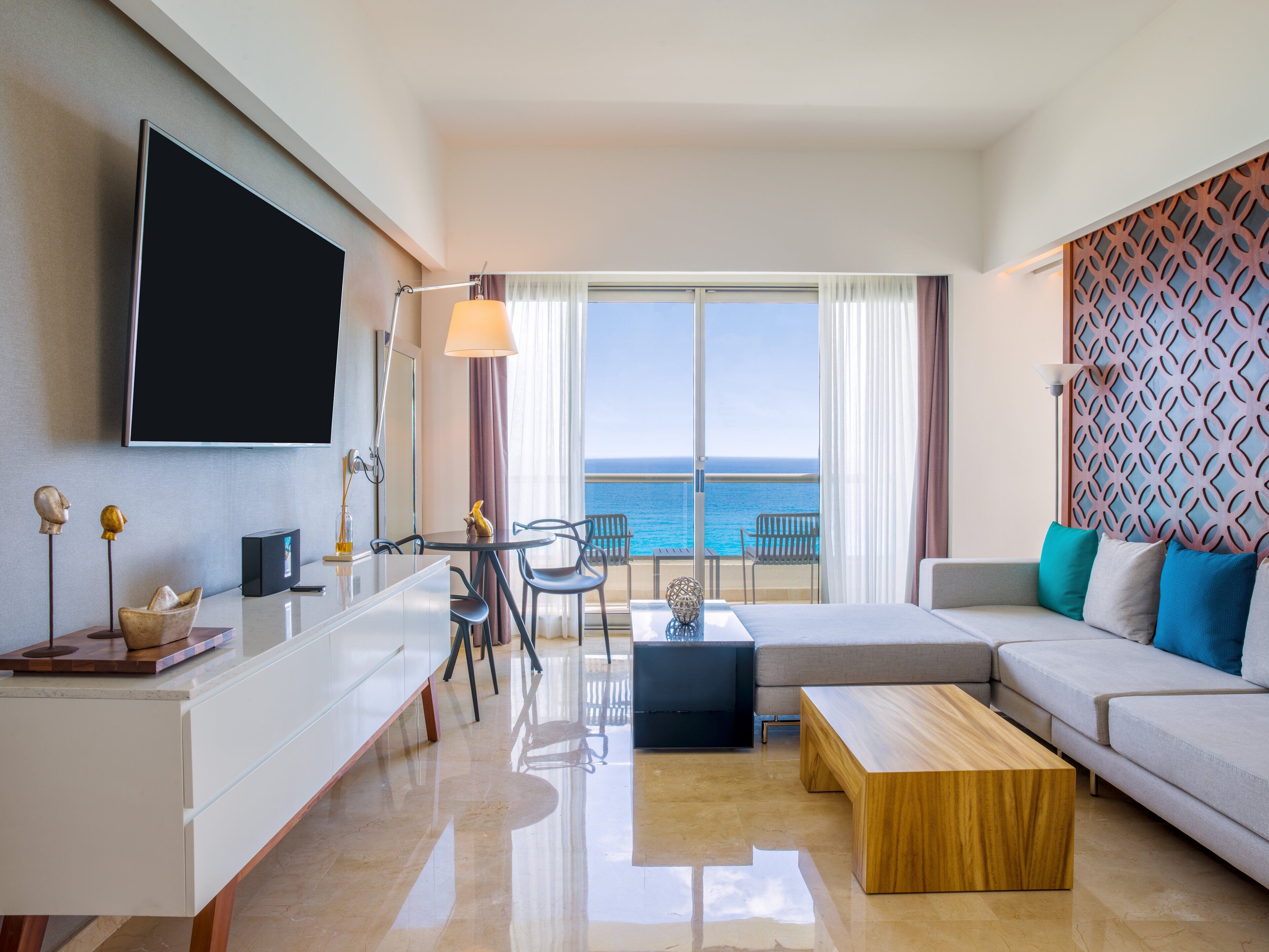 Quarto de hotel moderno com televisão grande, área de refeições, sofá e vista para o mar.