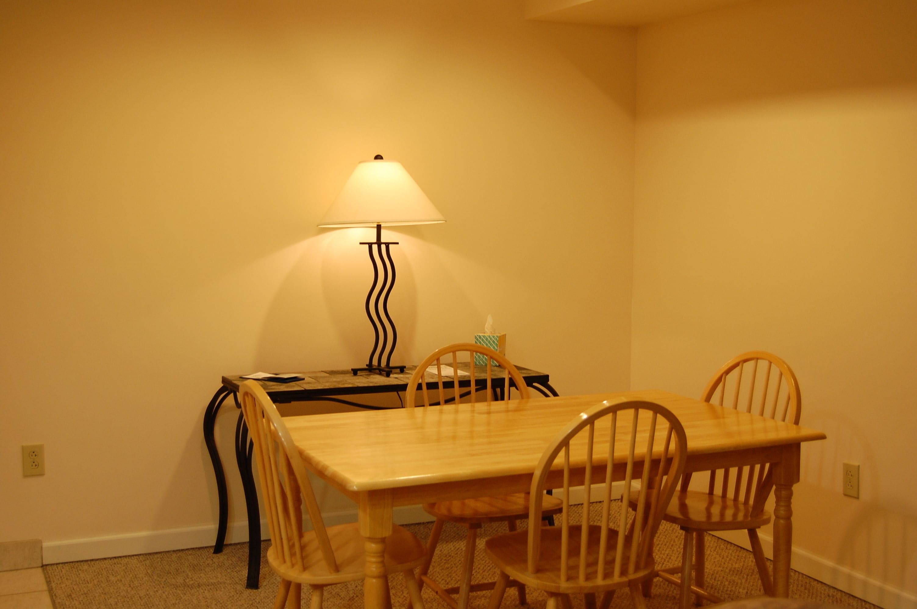 Uma área de refeições com mesa de madeira e quatro cadeiras, um abajur e uma mesinha com caixa de lenços.