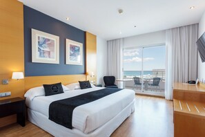 Quarto de hotel com uma cama grande, duas obras de arte emolduradas, uma escrivaninha com uma cadeira, uma pequena mesa com cadeiras e uma varanda com vista para o mar.