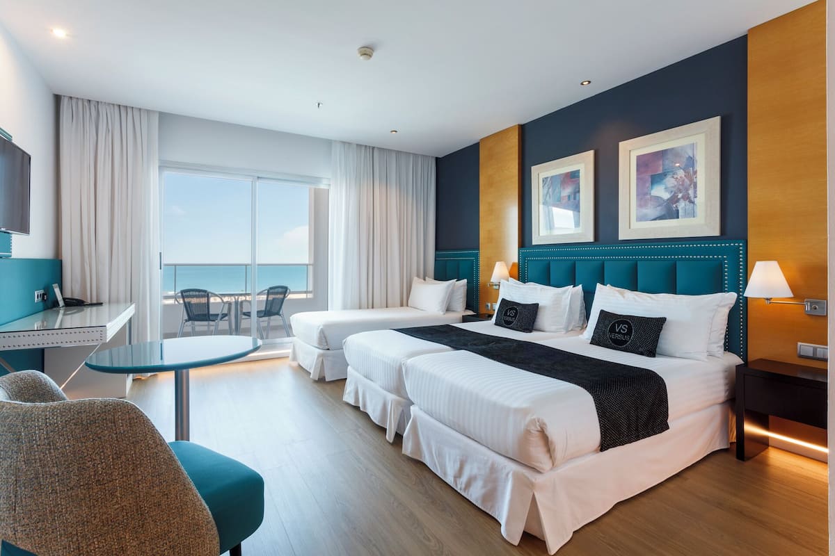 Quarto de hotel com duas camas, uma escrivaninha, uma cadeira e varanda com vista.