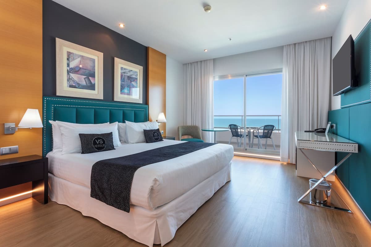 Quarto de hotel com uma cama grande, uma escrivaninha, uma televisão e varanda com vista para o mar.