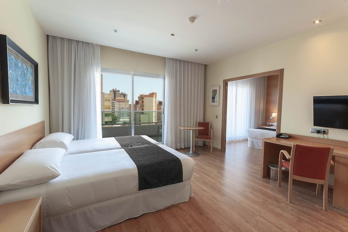 Quarto de hotel com duas camas, uma escrivaninha, uma cadeira, televisão e varanda com cortinas.