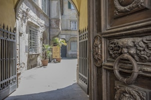Uma porta de madeira ornamentada com esculturas detalhadas, um vaso com planta e uma placa em um beco estreito, com paredes de pedra e piso de azulejos.