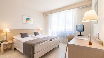 Quarto de hotel com cama, mesas de cabeceira, escrivaninha com computador e uma janela grande com cortinas.