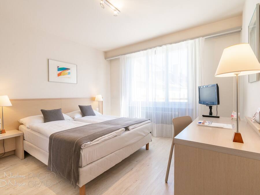 Quarto de hotel com cama, mesas de cabeceira, escrivaninha com computador e uma janela grande com cortinas.