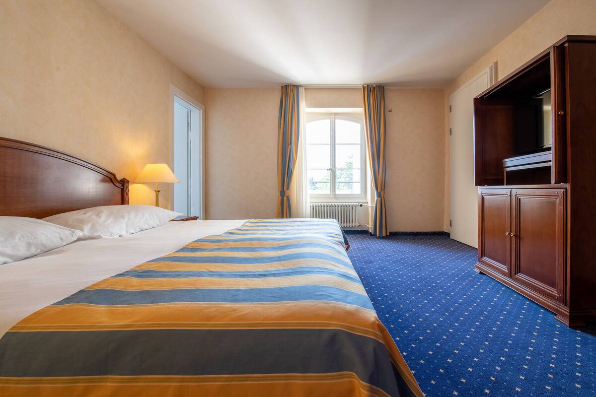 Quarto de hotel com uma cama grande, televisão, guarda-roupa e janela com cortinas.