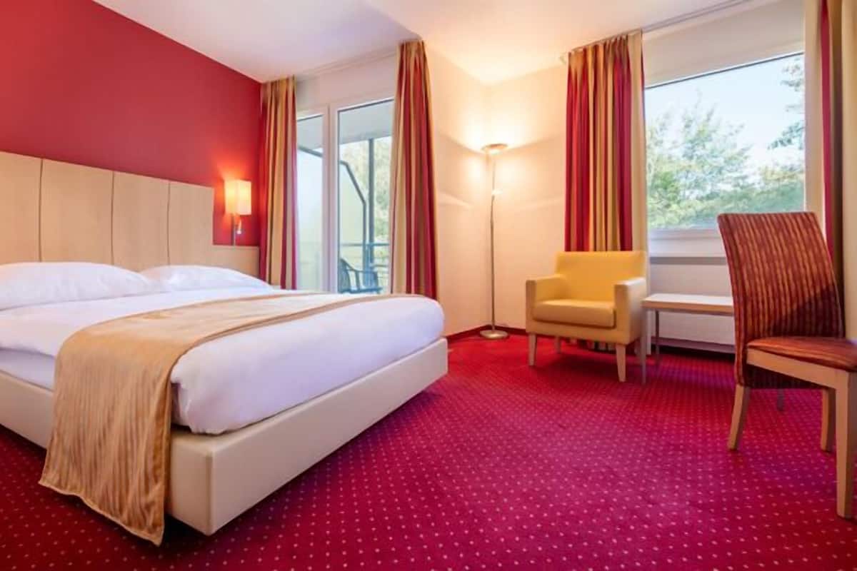 Quarto de hotel com uma cama grande, uma mesa, uma cadeira e uma janela com cortinas.