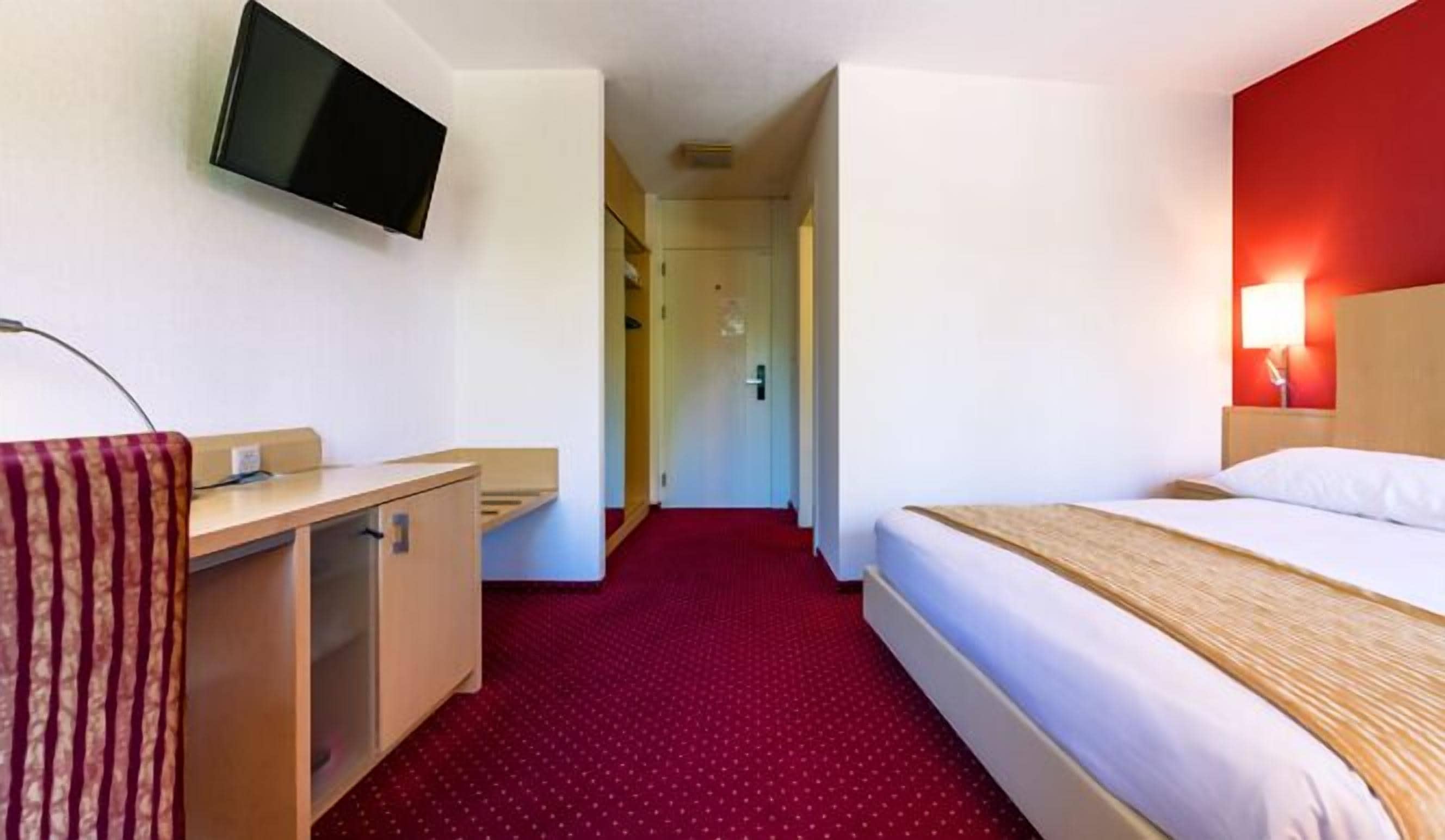Quarto de hotel com cama, escrivaninha, televisão fixada na parede e tapete vermelho.