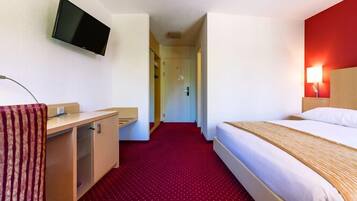 Quarto de hotel com cama, escrivaninha, televisão fixada na parede e tapete vermelho.