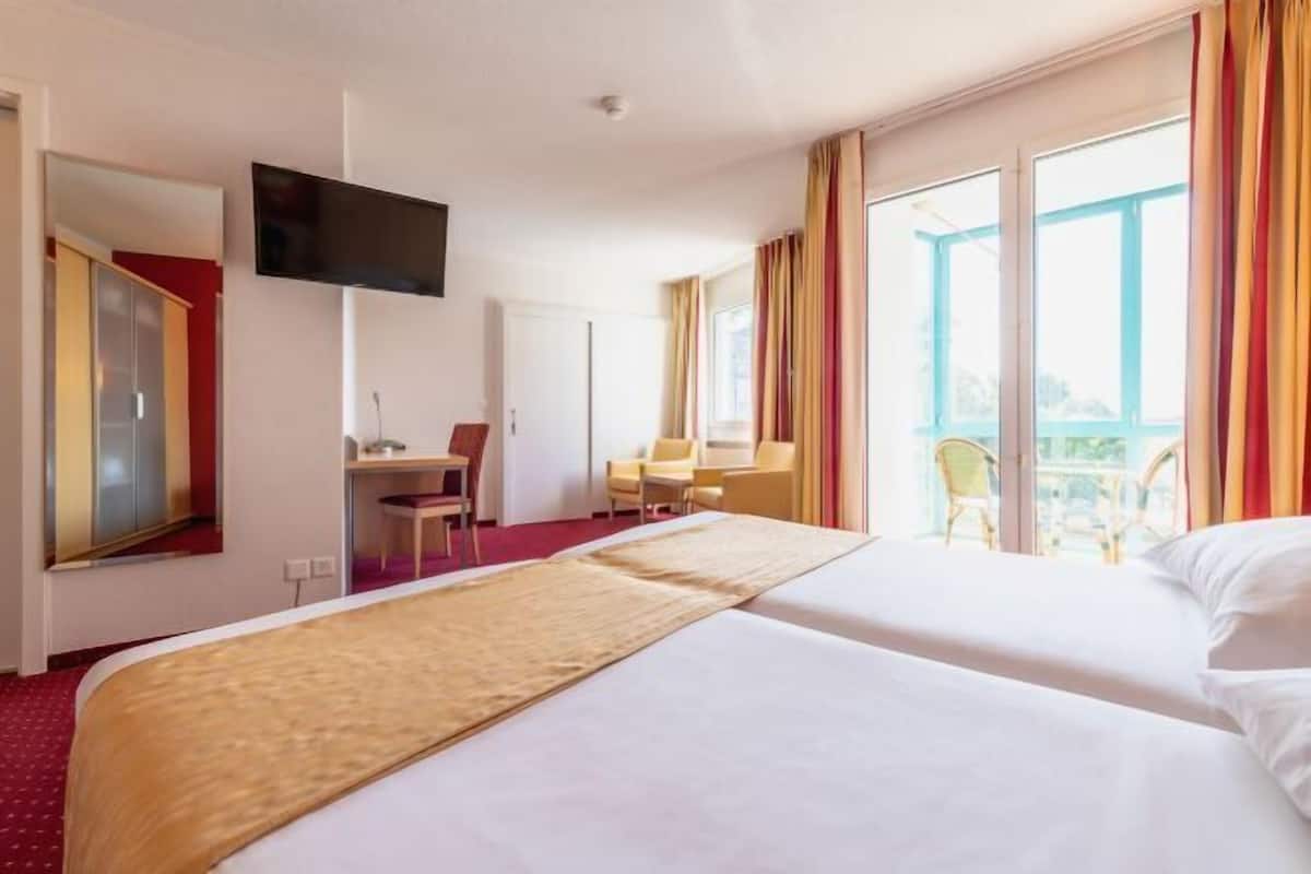 Quarto de hotel com duas camas, uma escrivaninha, uma cadeira, televisão e varanda com cortinas.